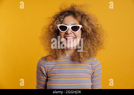 Happy young casual woman with fizzy hairstyle standing over yellow wall background, holding ...