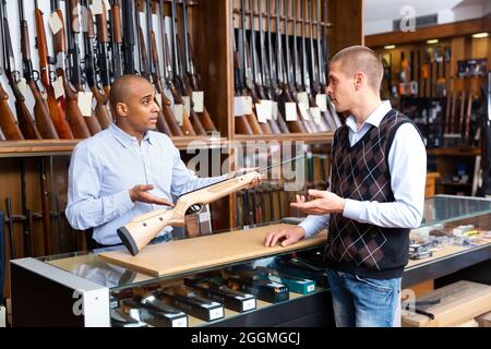 Latin american man owner of gun shop consulting customer about rifle ...