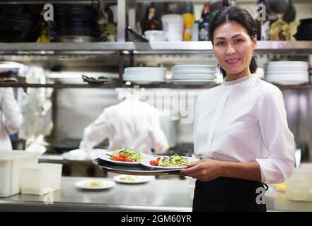 positive female waitstaff standing with tray in kitchen in restaurante ...