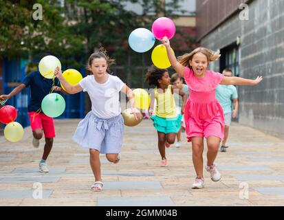Happy tweens wth colorful balloons chasing each other on city street ...