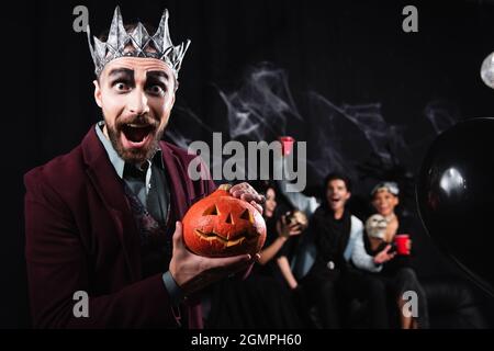 amazed man in vampire king halloween crown looking at camera on black ...