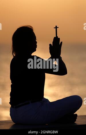 Silhouette of faithful woman praying with chrisrtian cross at sunset ...