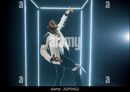 Stylish rapper in sunglasses poses in illuminated cube, dark background ...