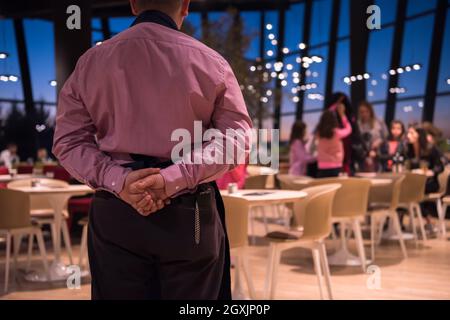 Rear view of young confident waiter standing with hands behind his back ...