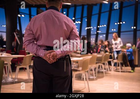 Rear view of young confident waiter standing with hands behind his back ...
