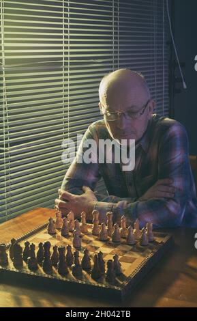 Old man sitting at table beside window with blinds and playing chess ...