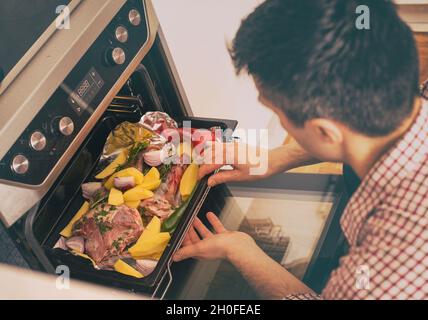 Satisfied young man putting casserole with meat and vegetables in oven ...