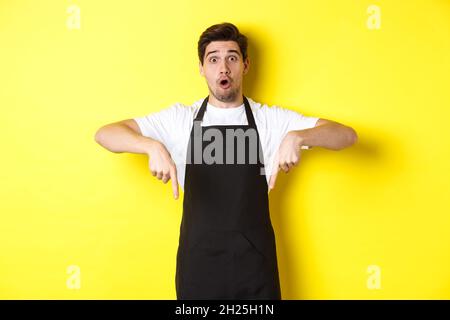 Surprised seller in black apron pointing fingers down, looking amazed ...