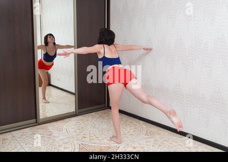A slender brunette does sports at the mirror in her apartment, standing ...