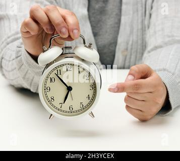 female hand holds a white metal alarm clock on a white table, time five ...