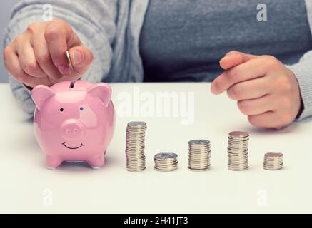 female hand throws a coin into a pink piggy bank on a white table ...