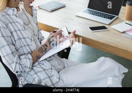 cropped view of tattooed woman writing in notebook near smartphone with ...