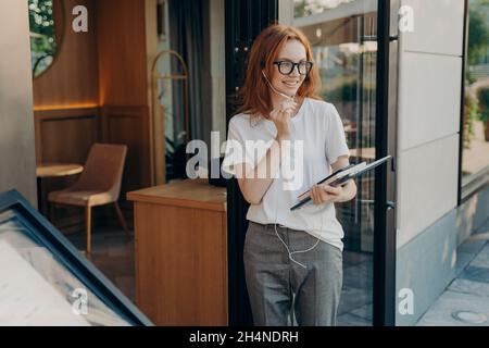 Outdoor shot of glad ginger young woman uses earphones with microphone ...