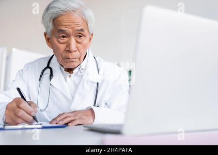 Elderly asian doctor writing on clipboard near blurred laptop in clinic ...