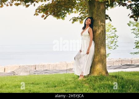 Smiling Colombian woman standing in white dress beneath leafy trees ...