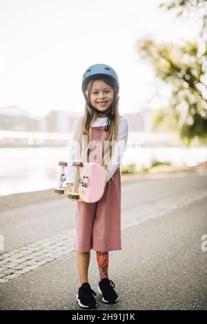 Smiling girl with skateboard standing in front of wall at footpath ...