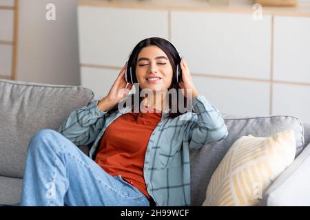 Young beautiful arab woman listening to music sitting on sofa at home ...
