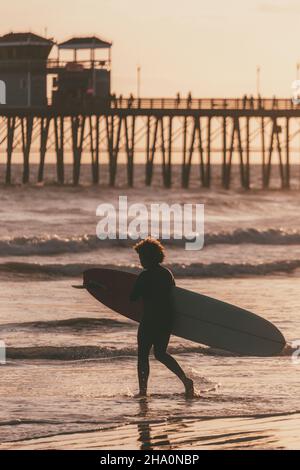 Woman carrying surfboard at sunset by pier Stock Photo - Alamy