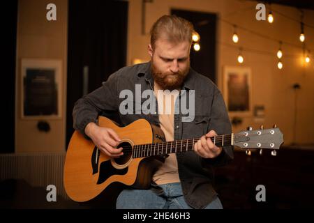 A young guy with a beard plays an acoustic guitar in a room with warm ...