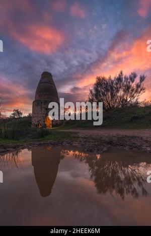 Aged lime kiln located on shore of calm reflective pond against cloudy ...