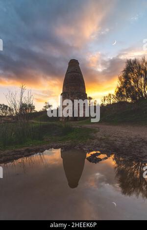 Aged lime kiln located on shore of calm reflective pond against cloudy ...