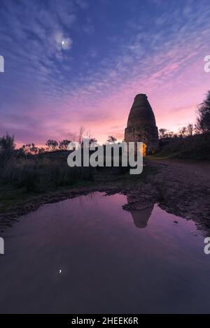Aged lime kiln located on shore of calm reflective pond against cloudy ...