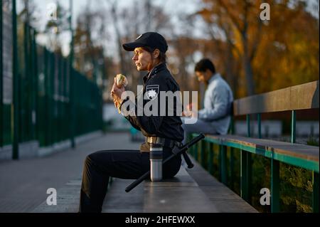 Hungry police woman eating sandwich sitting on park bench. Female cop ...