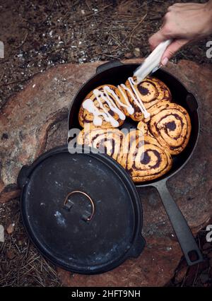 Hand pouring icing over cast iron made cinnamon rolls on a boulder ...