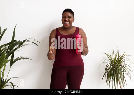 Sporty plus size woman with water bottle sitting on fitness mat at home ...