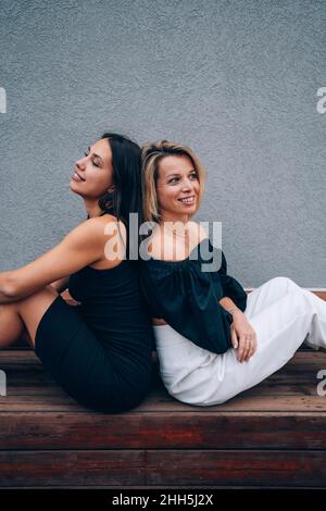 Two female friends sitting back to back on a bench outdoors enjoying ...