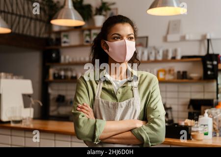 Young black waitress in face mask standing by cafe counter indoors ...