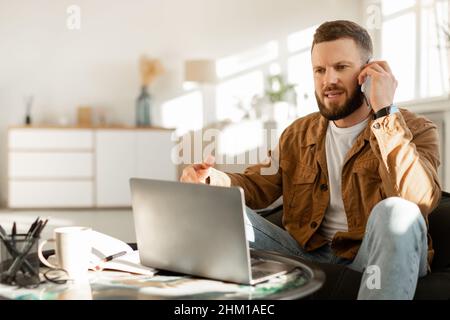 Displeased freelancer talking on smartphone near notebook and blurred ...
