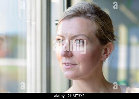 Quizzical blond woman with a knowing smile standing near a window with ...