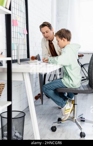 Preteen kid writing on sticky note on notebook near laptop at home ...
