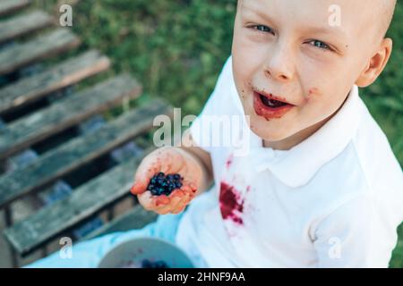 Pretty boy eating fresh berries with his hands and sitting on a wooden ...