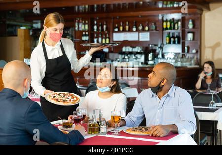 Waitress in mask serving cheerful people in pizzeria Stock Photo - Alamy