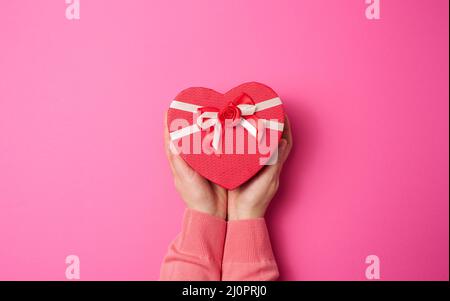 Two female hands hold a red heart-shaped box on a pink background ...