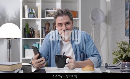50s man having breakfast checking news on phone drinking morning coffee ...