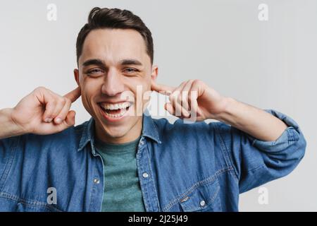 Young brunette man smiling and plugging his ears isolated over white ...