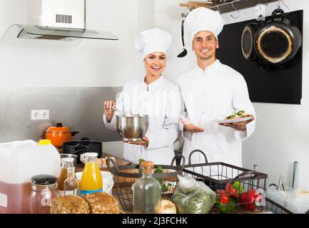 Two laughing female and male cooks wearing uniform working on kitchen ...