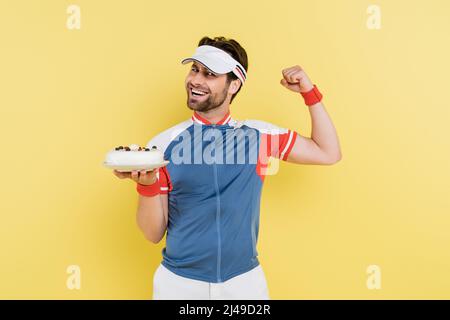 Cheerful sportsman holding cake and showing muscles isolated on yellow ...