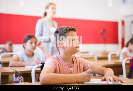 Tween boy listening to schoolteacher and writing in notebook in class ...
