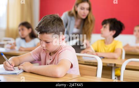Tween boy listening to schoolteacher and writing in notebook in class ...