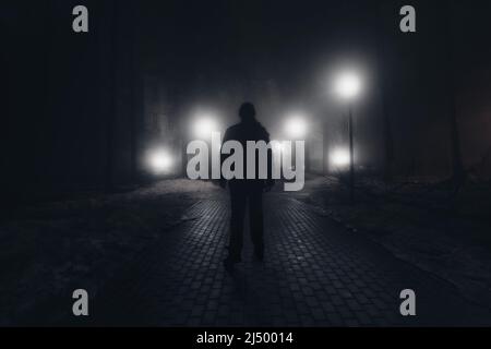 Sad man alone walking along the alley in night foggy park. Back view ...
