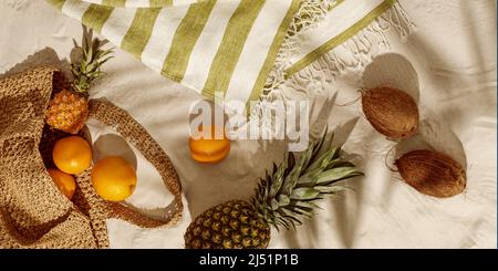 Fresh oranges and pineapple falling out from woven bag on sandy beach ...