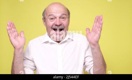Senior hispanic man in yellow tshirt pulling skin on face with hands ...