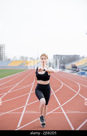 Smiling sportswoman with fitness tracker running on roof in evening ...