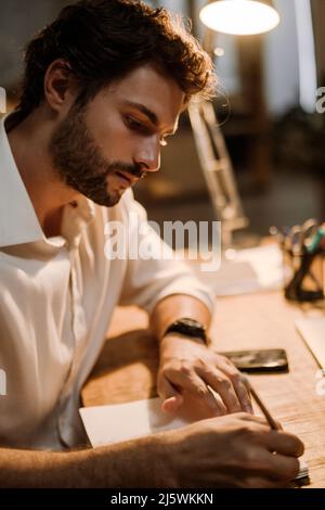 White bearded man writing down notes in planner while working at office ...