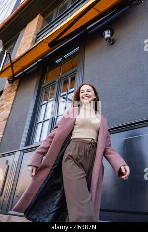 low angle view of joyful redhead woman in coat with scarf near modern ...