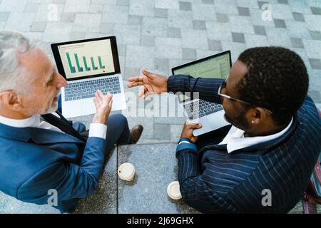 Multiracial men talking while working with laptops in cafe outdoors ...